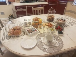 Uma mesa de café da manhã posta com pães, frutas e bebidas, incluindo um copo de suco de laranja e uma garrafa de leite de soja.