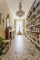 Uma biblioteca com chão em padrão hexagonal, um candelabro, estantes de livros, uma cadeira de madeira e uma mesa com abajur.