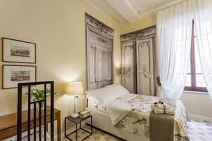 Quarto com cama, mesa de cabeceira, abajur, cadeira e duas fotos emolduradas na parede.