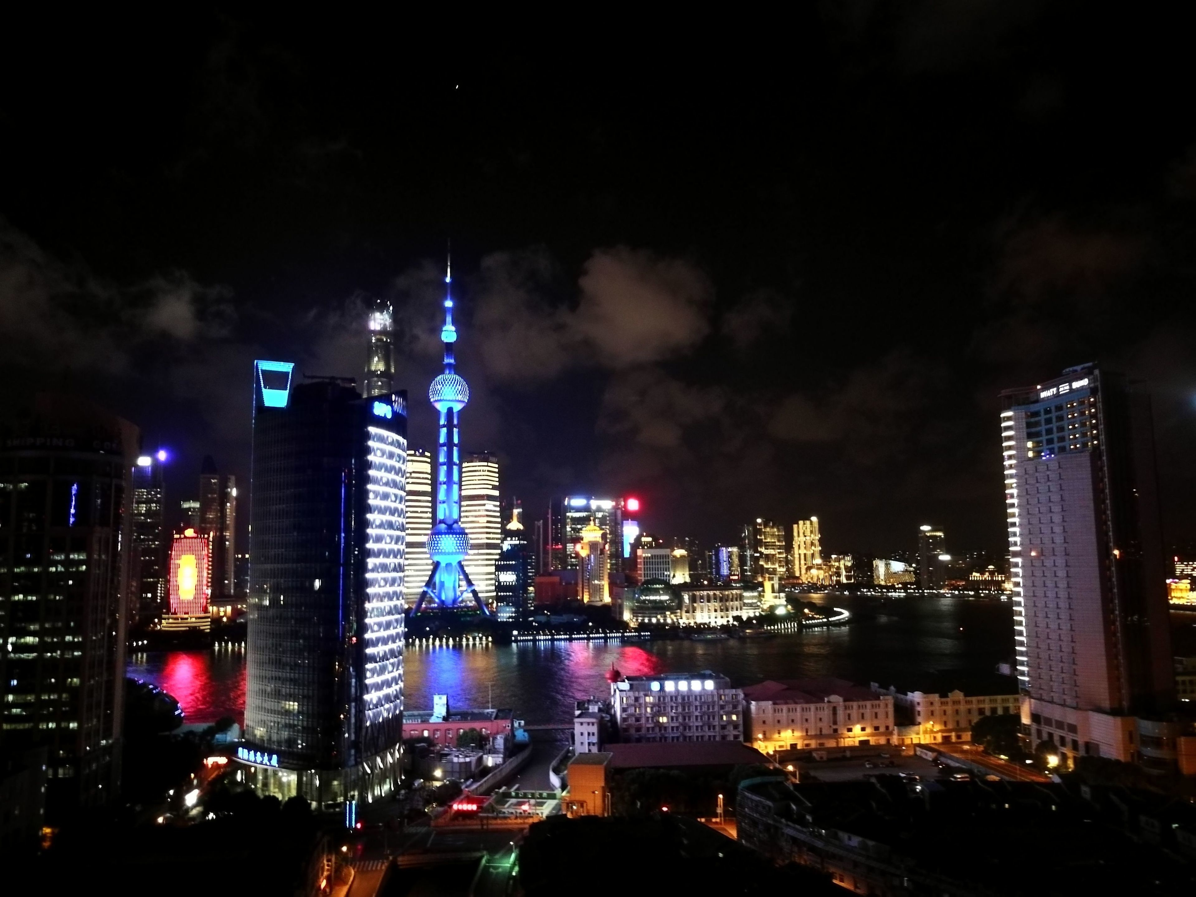 Uma paisagem urbana noturna com edifícios iluminados e uma torre imponente com uma agulha iluminada em azul.
