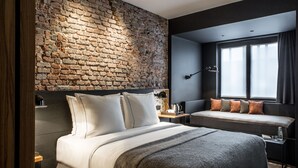 Quarto moderno com parede de tijolos, uma cama grande com várias almofadas, uma mesinha de cabeceira, um sofá pequeno com almofadas e uma janela com cortinas.