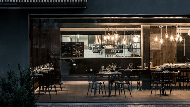 Uma área de refeições ao ar livre com mesas e cadeiras, um bar com balcão e luminárias pendentes.
