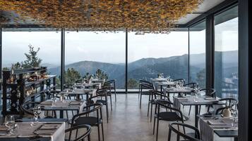Um restaurante com mesas preparadas para refeições, amplas janelas com vista para montanhas e um teto decorado com folhas penduradas.