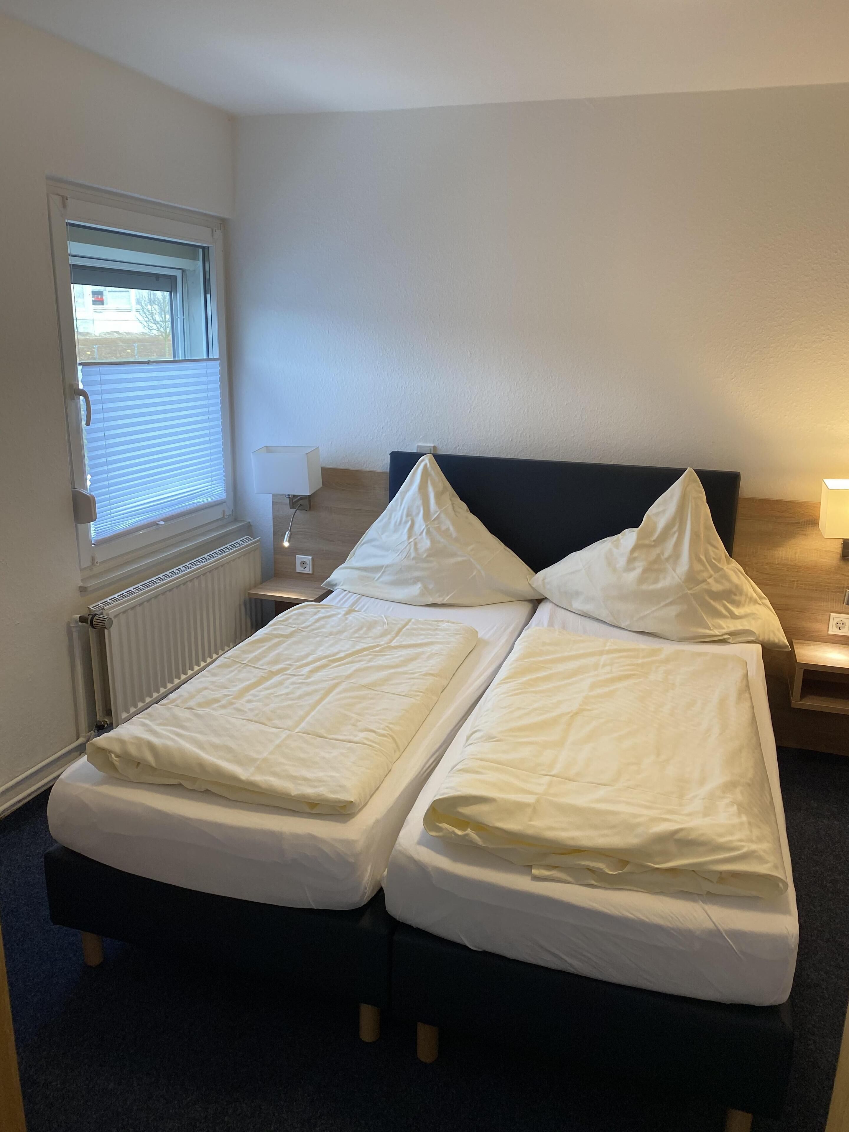 Uma cama de casal com cabeceira preta, uma janela com persianas, uma mesa de cabeceira com abajur e um radiador.