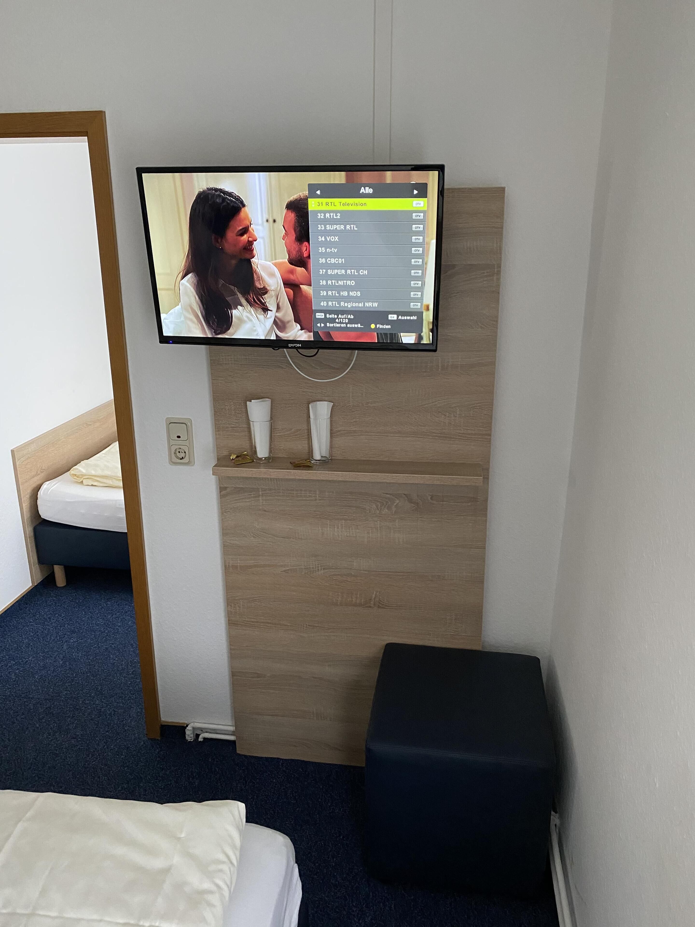 Quarto de hotel com televisão presa na parede exibindo uma lista de canais.