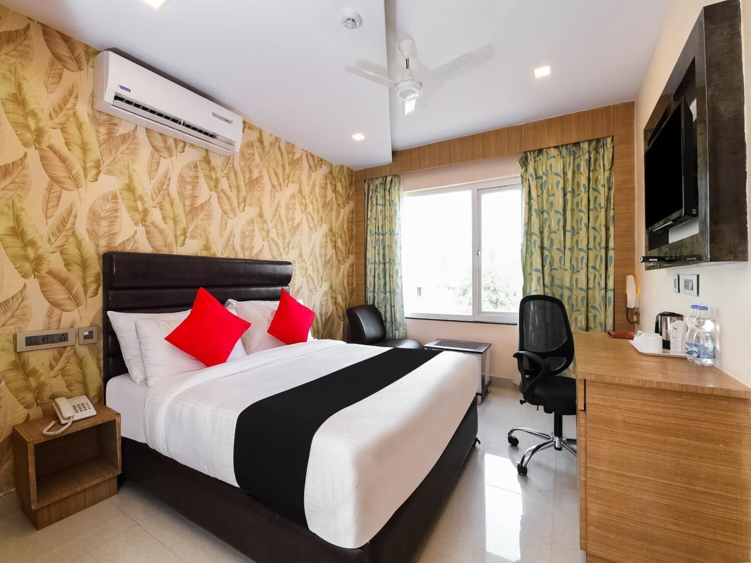Quarto de hotel com cama, escrivaninha, cadeira, ar condicionado, ventoinha no teto, janela com cortinas e televisão.