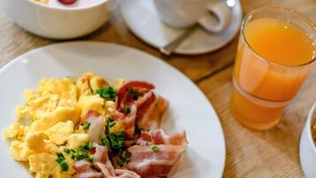 Prato de café da manhã com ovos mexidos, bacon e cebolinha, um copo de suco de laranja e uma xícara de café sobre uma mesa de madeira.