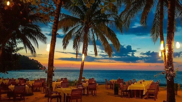 Um restaurante à beira-mar com mesas e cadeiras dispostas para refeições, palmeiras e vista para o oceano ao pôr do sol.