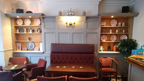 Uma área de refeições com mesa de madeira e cadeiras estofadas, cercada por prateleiras com pratos decorativos e vasos.