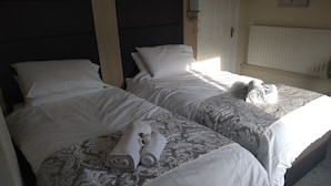 Duas camas de solteiro com roupa de cama branca e colchas estampadas, uma toalha enrolada em cada cama e uma luminária fixa na parede.