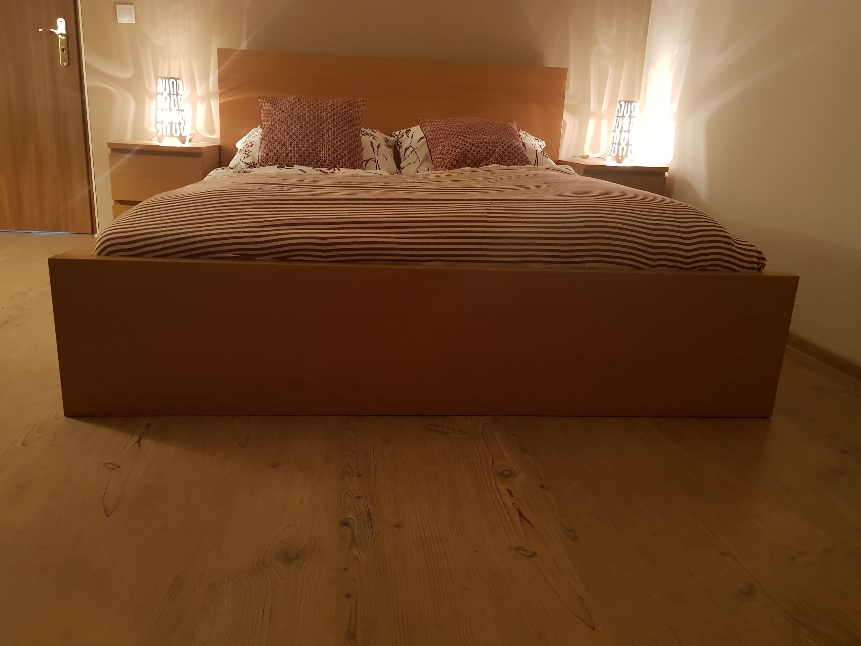Uma cama grande e retangular com cabeceira de madeira e roupa de cama listrada. Há duas mesas de cabeceira com luminárias de cada lado da cama.