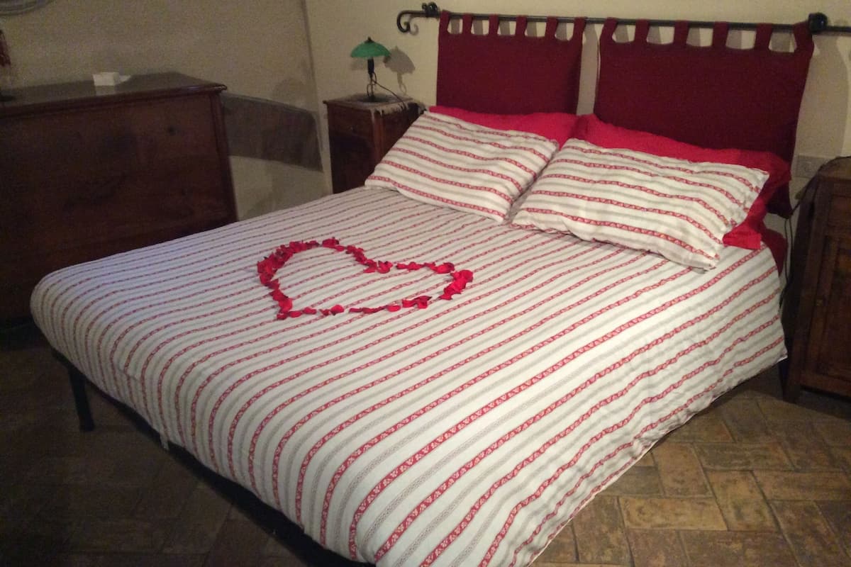 Uma cama com uma decoração em formato de coração na colcha, um cabeceira vermelha e uma mesa de cabeceira de madeira com uma luminária.