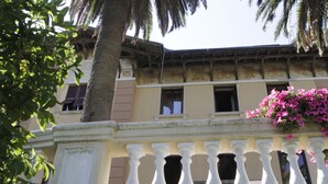 Um edifício com varanda, palmeiras e flores cor-de-rosa.