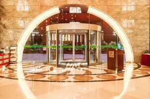 Entrada de hotel com porta giratória, piso de mármore e tapete vermelho que leva à área de recepção.