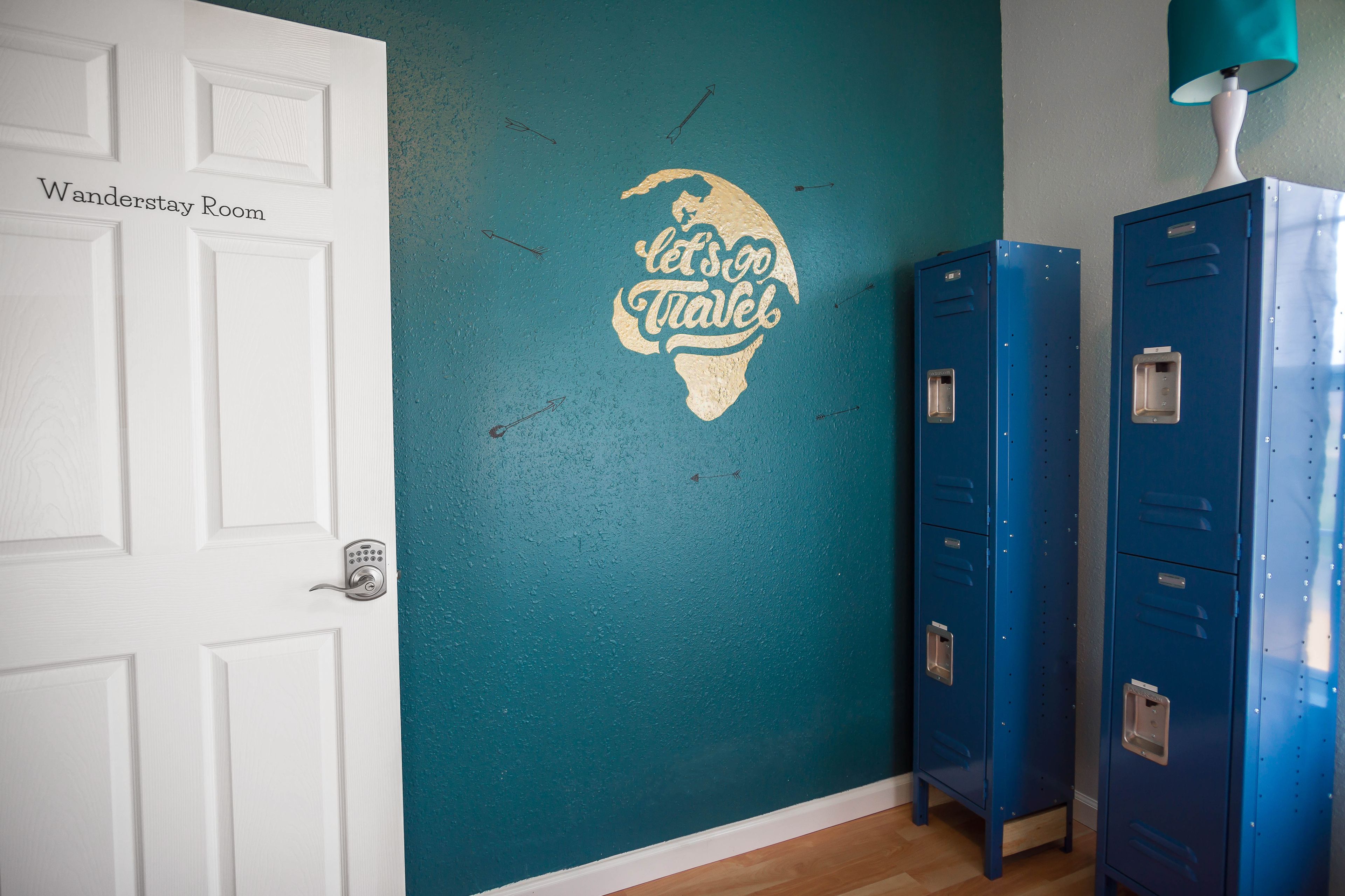 Quarto com parede verde-água, porta branca com a inscrição 'Wanderstay Room' e armários azuis.