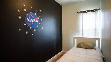 Quarto com adesivo de ônibus espacial da NASA, uma cama com travesseiro bege e uma janela com cortinas.