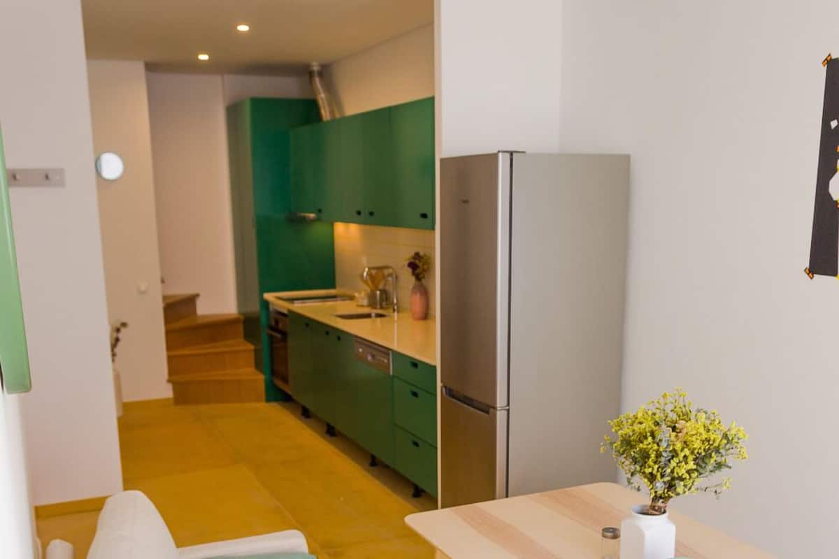 Cozinha moderna com armários verdes, geladeira de aço inoxidável e área de jantar com mesa e banco.