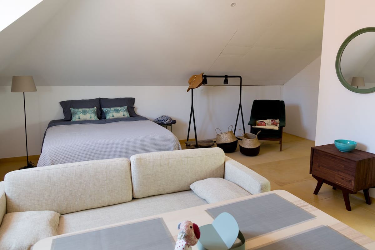 Quarto com cama, sofá, mesa pequena, espelho e luminária suspensa.