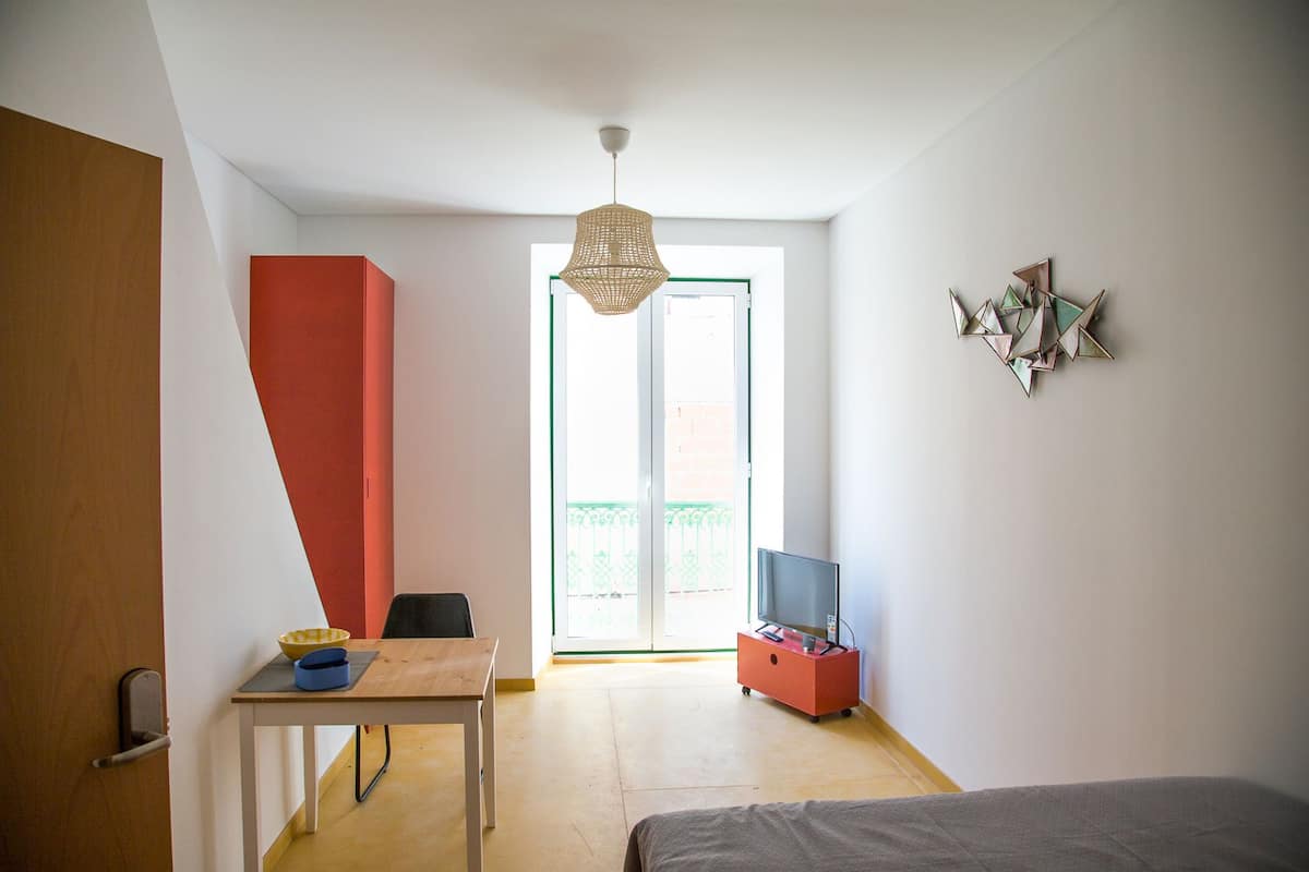 Quarto com cama, escrivaninha, cadeira, televisão sobre suporte, porta e janela com vista externa.
