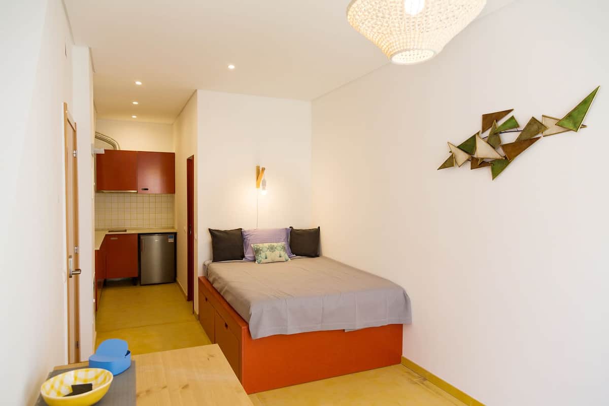 Quarto compacto com cama, mesa de cabeceira e um pequeno banquinho. Anexo, uma kitchenette com frigorífico, fogão e armários. Uma peça decorativa na parede é visível.