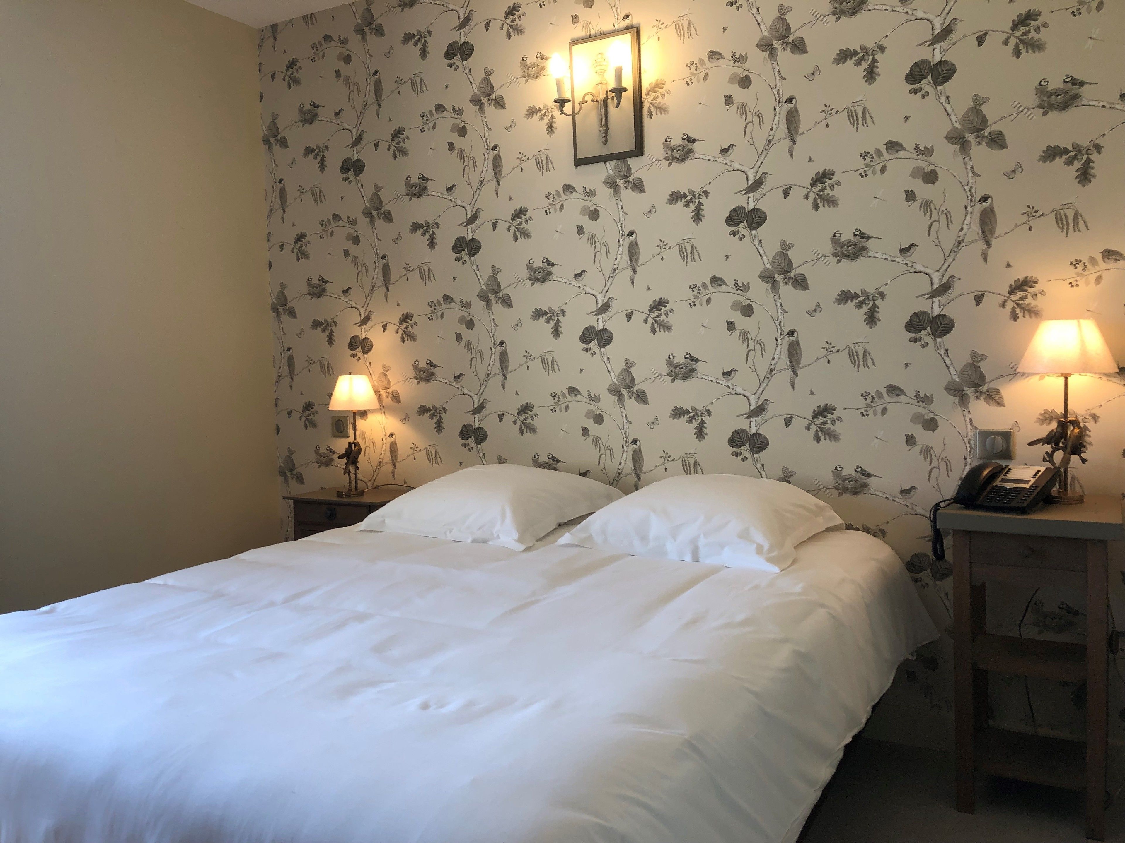 Cama bem arrumada com lençóis brancos, duas luminárias laterais e um telefone num criado-mudo de madeira.