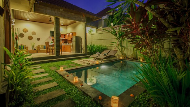 Piscina com degraus de pedra, rodeada por vegetação e área externa bem iluminada, com uma espreguiçadeira e uma mesa de jantar.