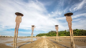Uma trilha leva a uma praia, onde se destacam três estruturas altas e esguias que lembram lanternas tradicionais.
