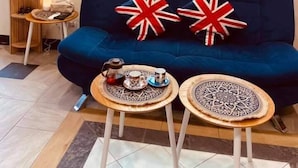 Um sofá azul com duas almofadas, duas mesas redondas de madeira com pratos decorativos, uma pequena estante de madeira com um urso de pelúcia e um cartaz na parede.