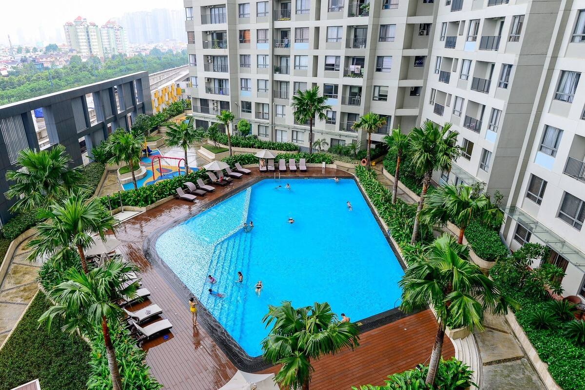 Uma piscina no terraço rodeada por palmeiras e espreguiçadeiras, com vista para edifícios altos ao fundo.