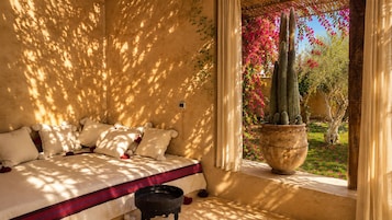 Quarto com cama, um cacto grande em vaso e vista para vegetação e flores.