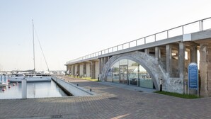 Uma orla com uma passarela e uma ponte, um porto com barcos e um edifício de vidro com arco.