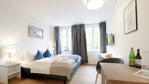 Quarto de hotel com cama, escrivaninha, cadeira, mesa de cabeceira, abajur, janela com cortinas e um quadro na parede.