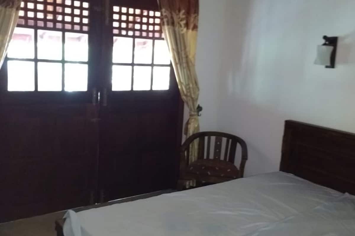 Quarto com cama, cadeira de madeira, janela com cortinas e luminária na parede.