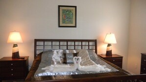 Cama bem arrumada com arranjo de toalhas em formato de coração, duas luminárias de cabeceira e uma obra de arte emoldurada na parede.