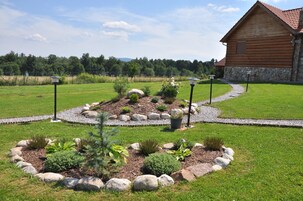 Uma cabana de toras com base de pedra, cercada por um jardim bem cuidado com um caminho de cascalho, arbustos variados e alguns vasos de plantas.