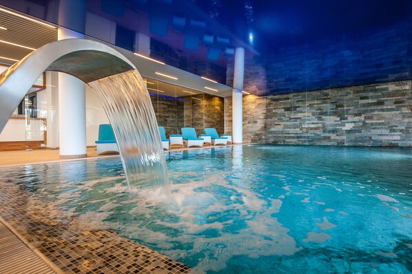Piscina interior com efeito cascata, cadeiras de descanso azuis e parede de pedra ao fundo.