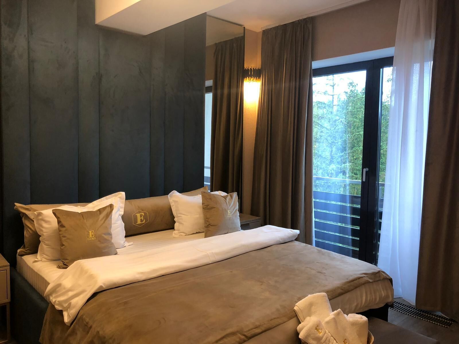 Quarto de hotel moderno com uma cama grande, paredes escuras e uma varanda com cortinas.