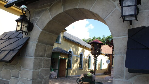 Um arco com paredes de pedra, um carrinho de madeira com flores e um edifício com telhado vermelho ao fundo.
