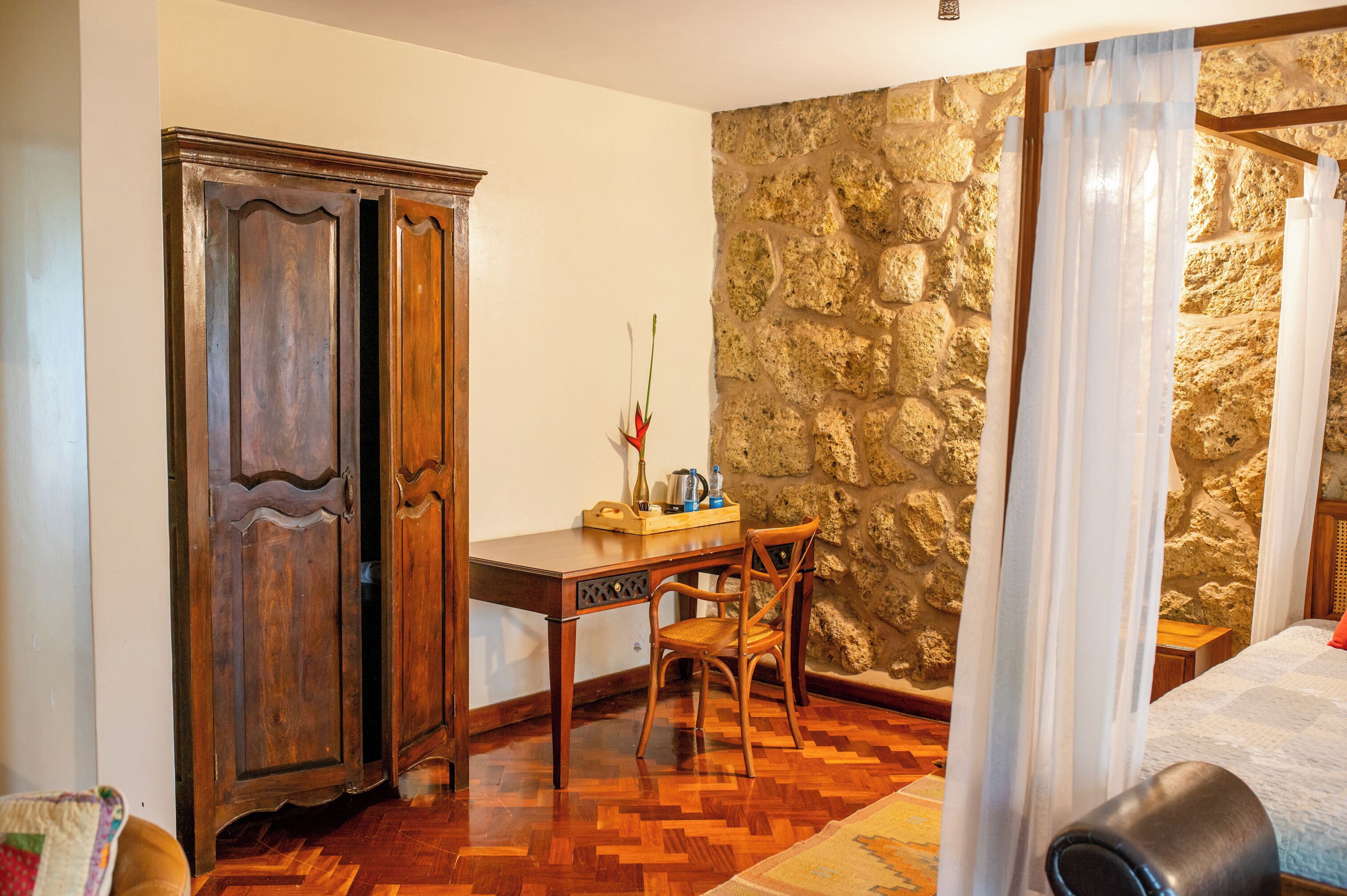Quarto com guarda-roupa de madeira, escrivaninha com cadeira, parede de pedra e cama com dossel.