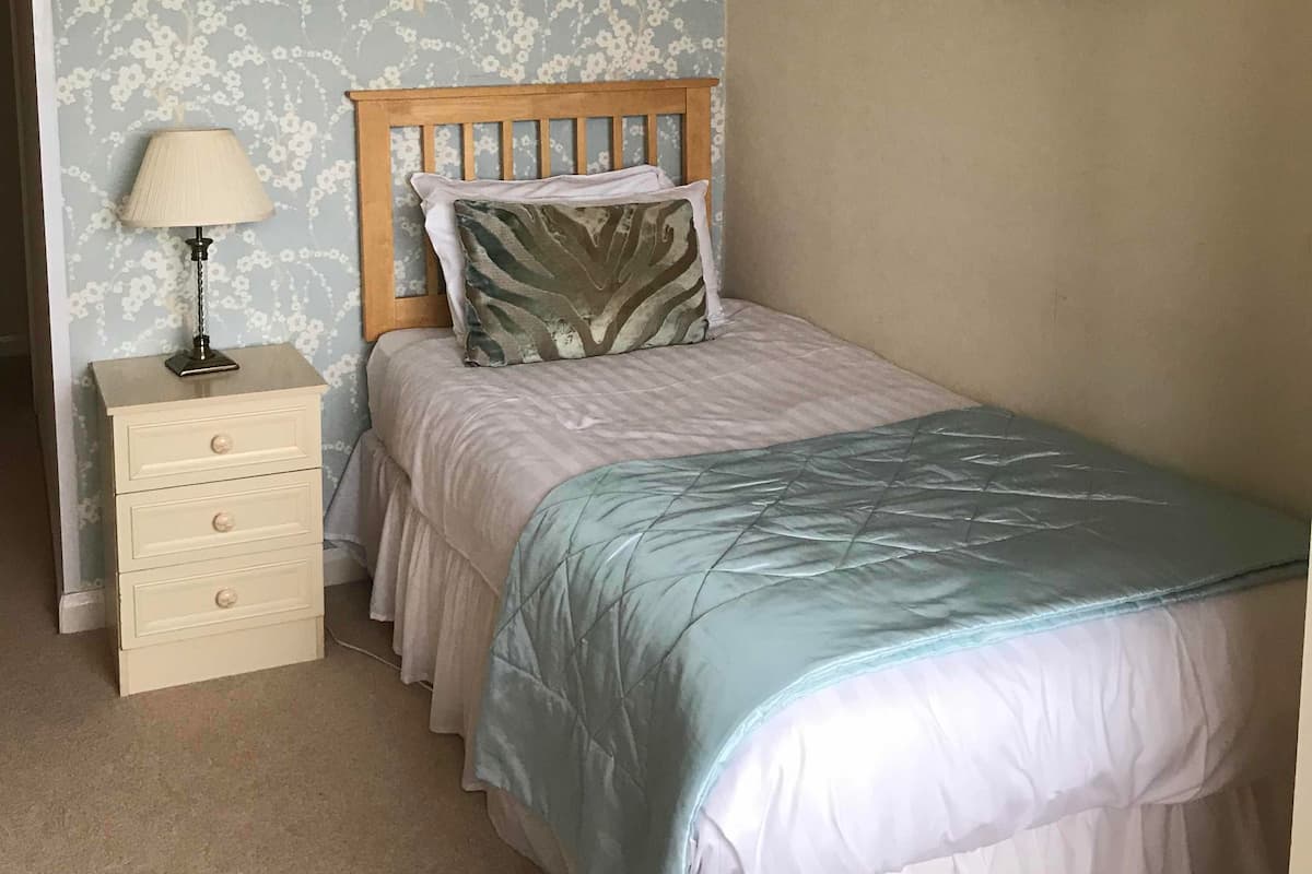 Quarto com cama, mesa de cabeceira, abajur, quadro na parede e papel de parede com padrão floral.