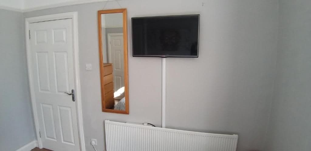 Quarto com porta branca, espelho, televisão na parede e radiador.