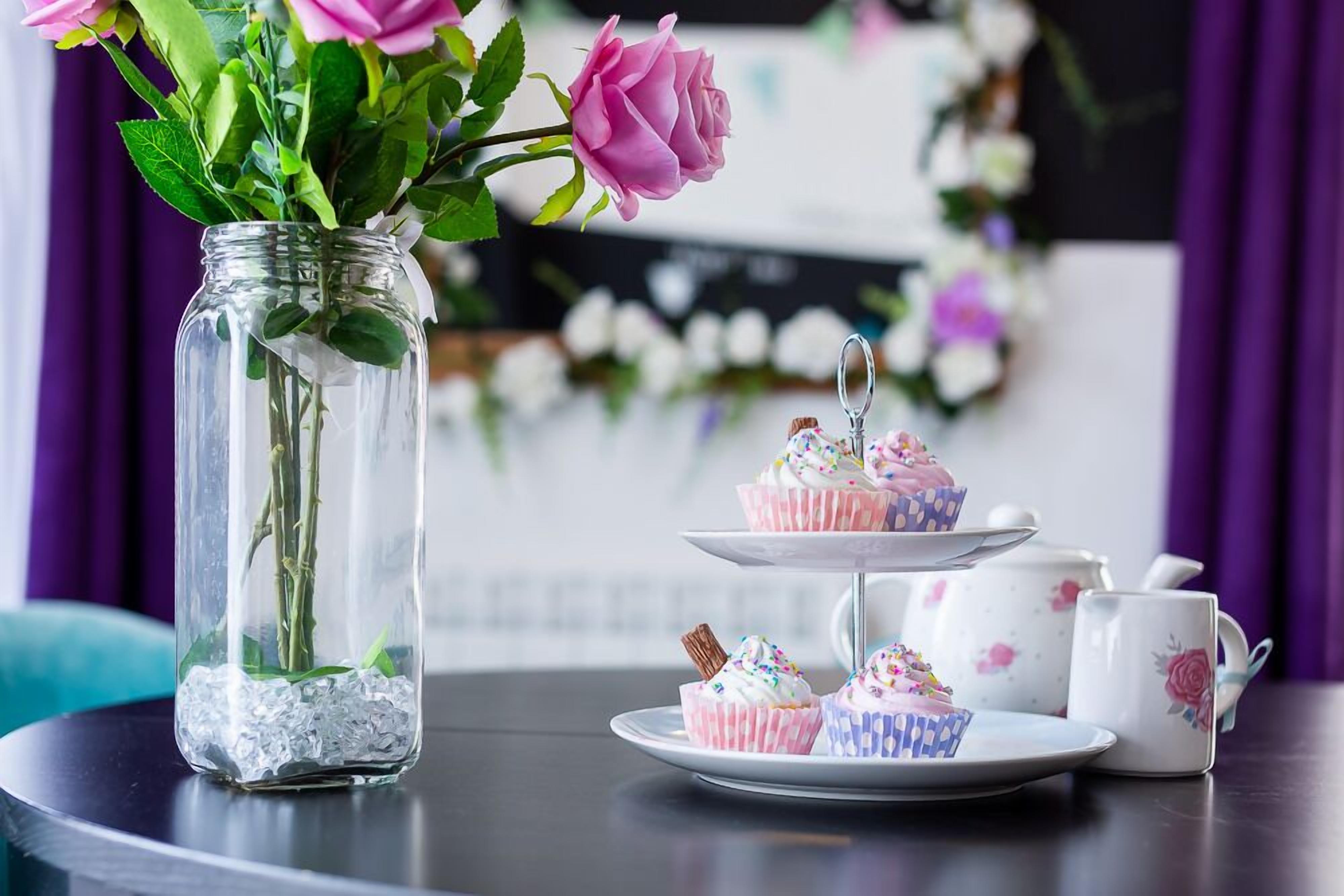 Uma mesa com um frasco de vidro contendo uma rosa, um suporte escalonado com cupcakes e um conjunto de chá.