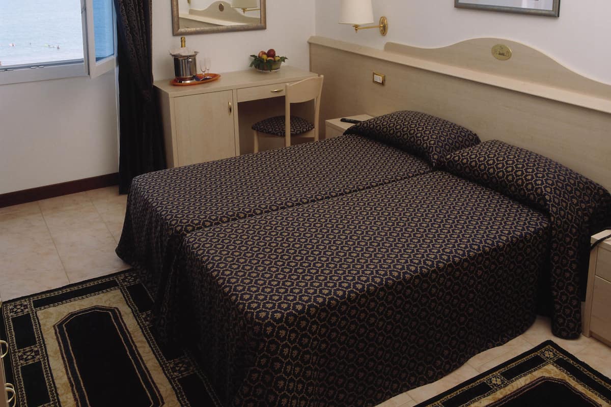 Quarto de hotel com cama, escrivaninha, cadeira, mesa de cabeceira, abajur, quadro emoldurado e janela com cortinas.