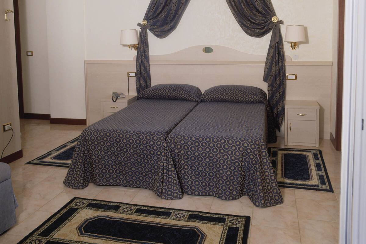 Quarto com cama de casal, duas mesas de cabeceira, um criado-mudo e uma cama com dossel, além de um tapete estampado.