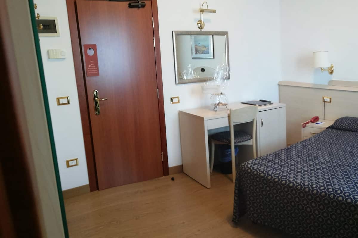 Quarto de hotel com cama, escrivaninha, cadeira, guarda-roupa, espelho, mesa de cabeceira e criado-mudo.