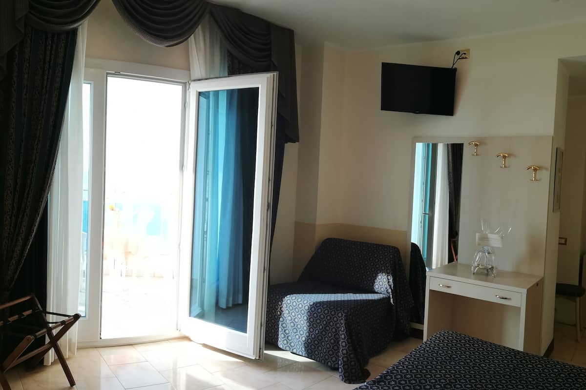 Quarto de hotel com cama, escrivaninha, cadeira, televisão e porta de correr para a varanda.