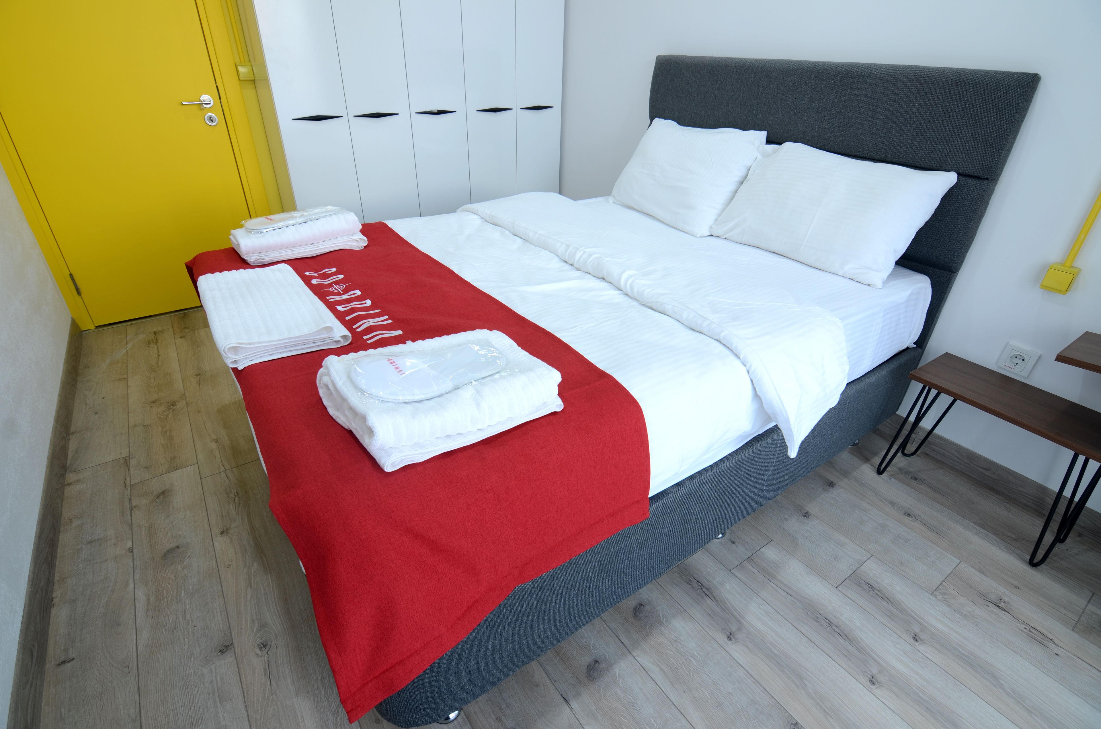 Uma cama bem arrumada com lençóis brancos e um cobertor vermelho, uma mesa de cabeceira de madeira com uma luminária, e uma porta amarela.