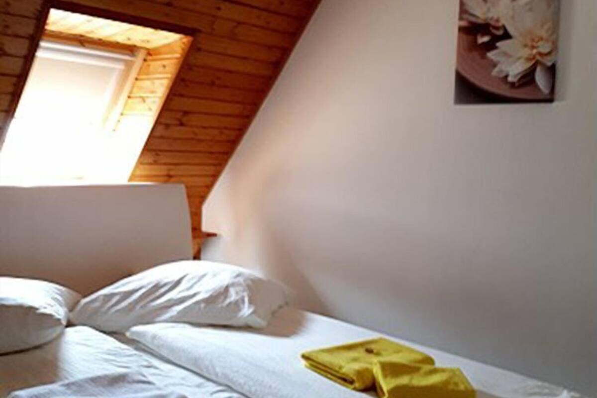 Quarto com teto inclinado, uma cama com roupa de cama branca, uma toalha amarela e um quadro de flores na parede.