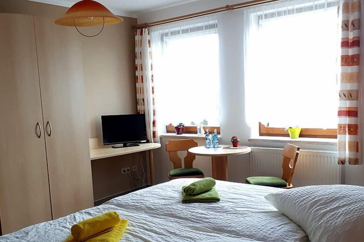 Quarto de hotel com cama, escrivaninha com televisão, pequena mesa com cadeiras e janela com cortinas.