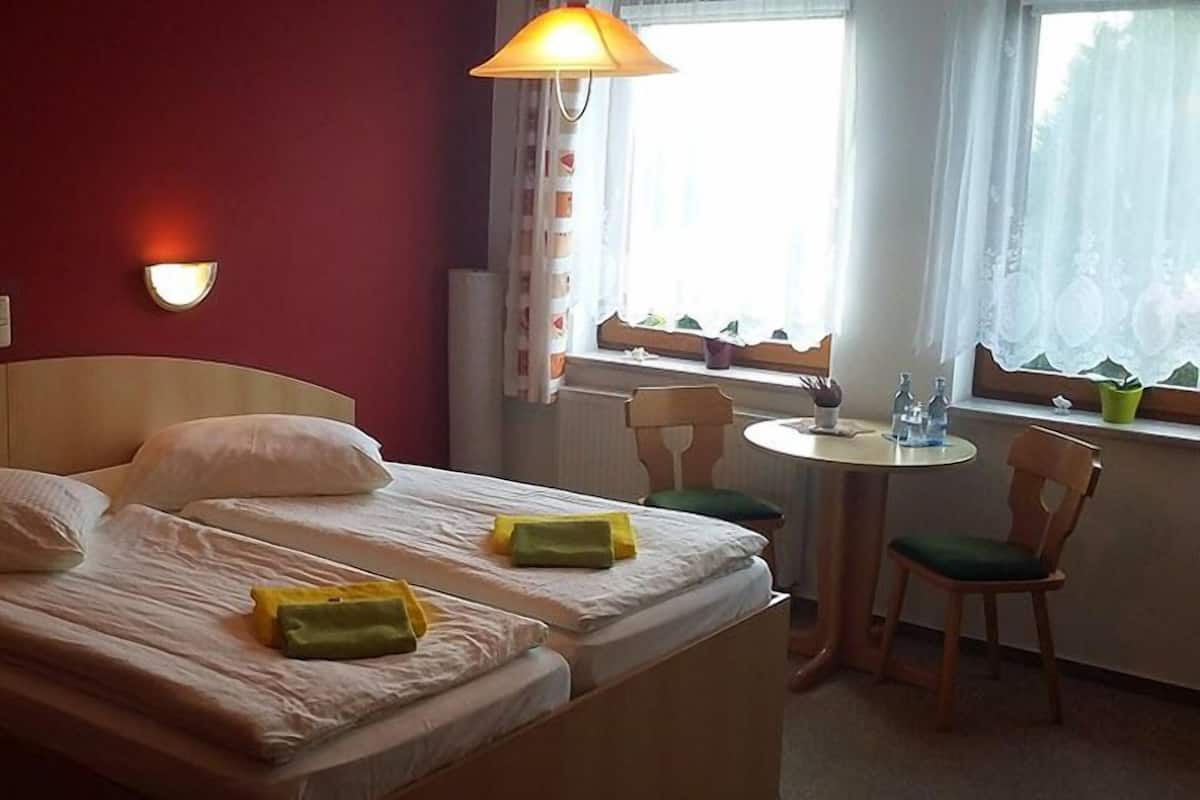 Quarto de hotel com uma cama, duas luminárias de cabeceira, uma mesinha com uma cadeira e uma janela com cortinas.